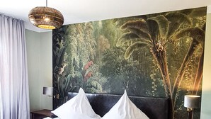 Cama bem arrumada com lençóis e almofadas brancas, cabeceira escura e um grande mural tropical na parede ao fundo.