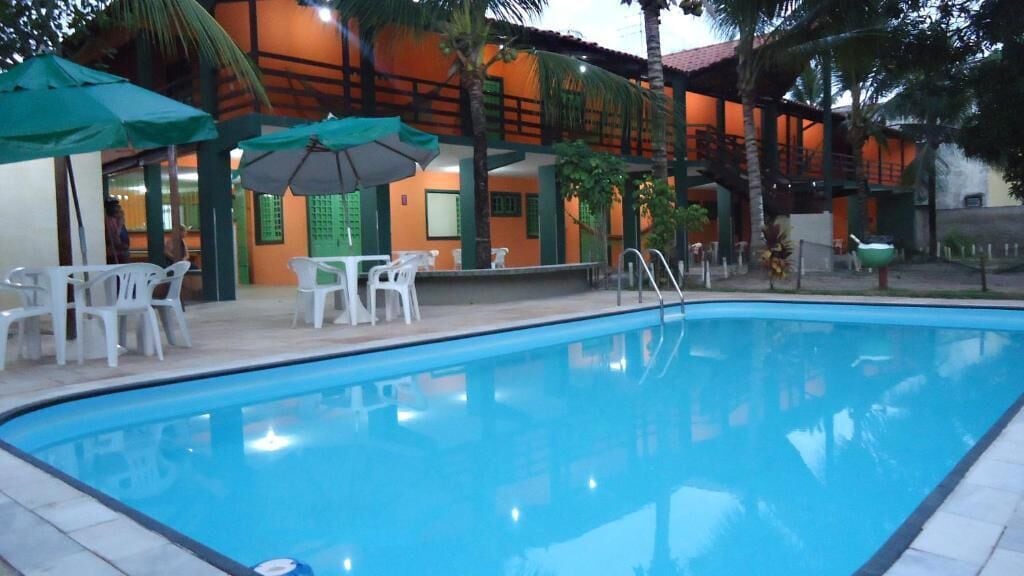 Uma piscina com cadeiras e guarda-sóis brancos, cercada por um edifício com paredes laranja e detalhes verdes.