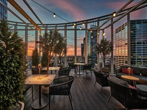 Terraço no telhado com mesas, cadeiras e plantas em vasos, com vista para a paisagem urbana ao pôr do sol.
