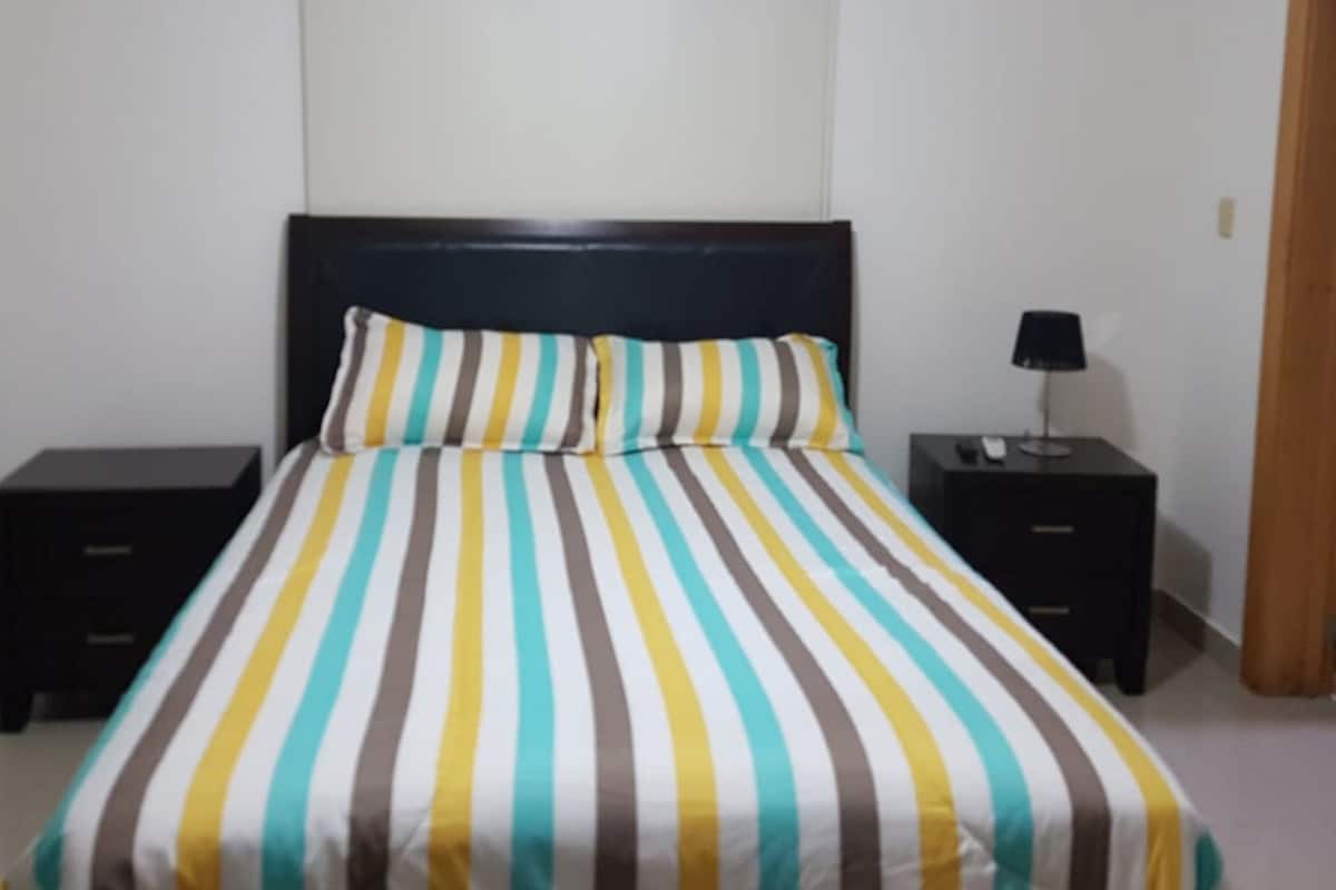 Quarto com colcha listrada, cabeceira escura, duas mesas de cabeceira, abajur e janela com persianas.