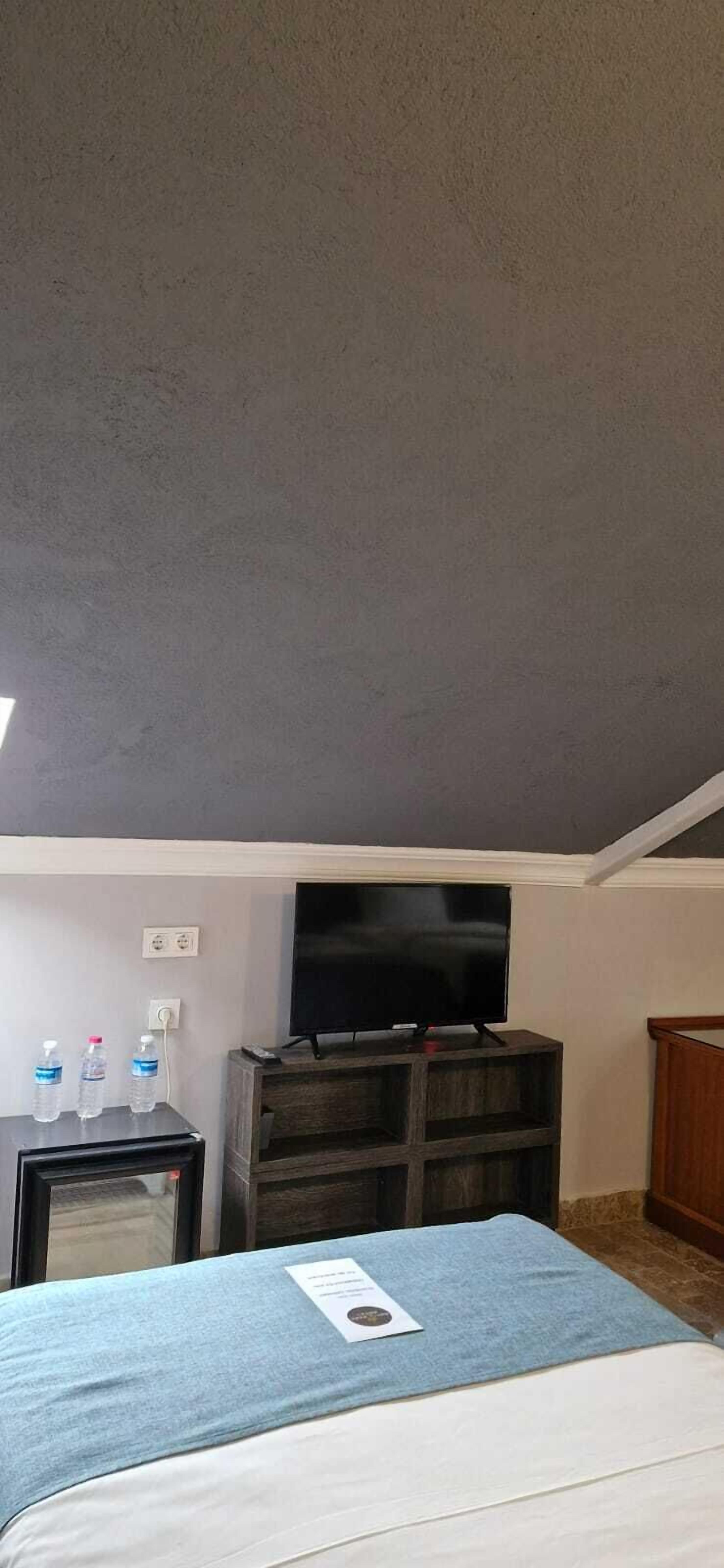 Quarto de hotel com cama, televisão sobre suporte de madeira e garrafa de água em uma mesinha.