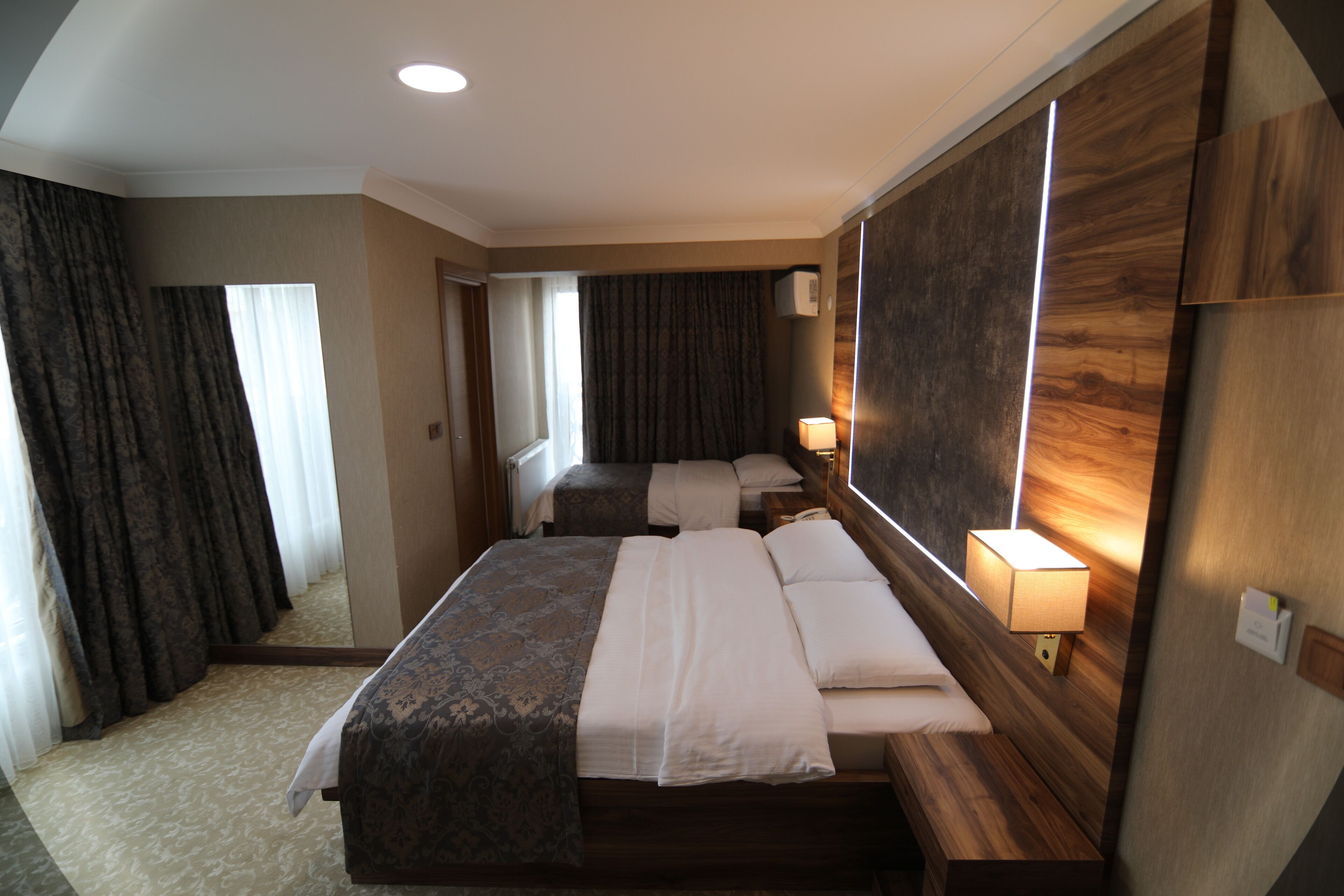 Quarto de hotel com uma cama grande, uma mesinha de cabeceira com abajur, um espelho e vista para o corredor.