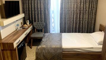 Quarto de hotel com cama, escrivaninha com televisão, cadeira e janela com cortinas.
