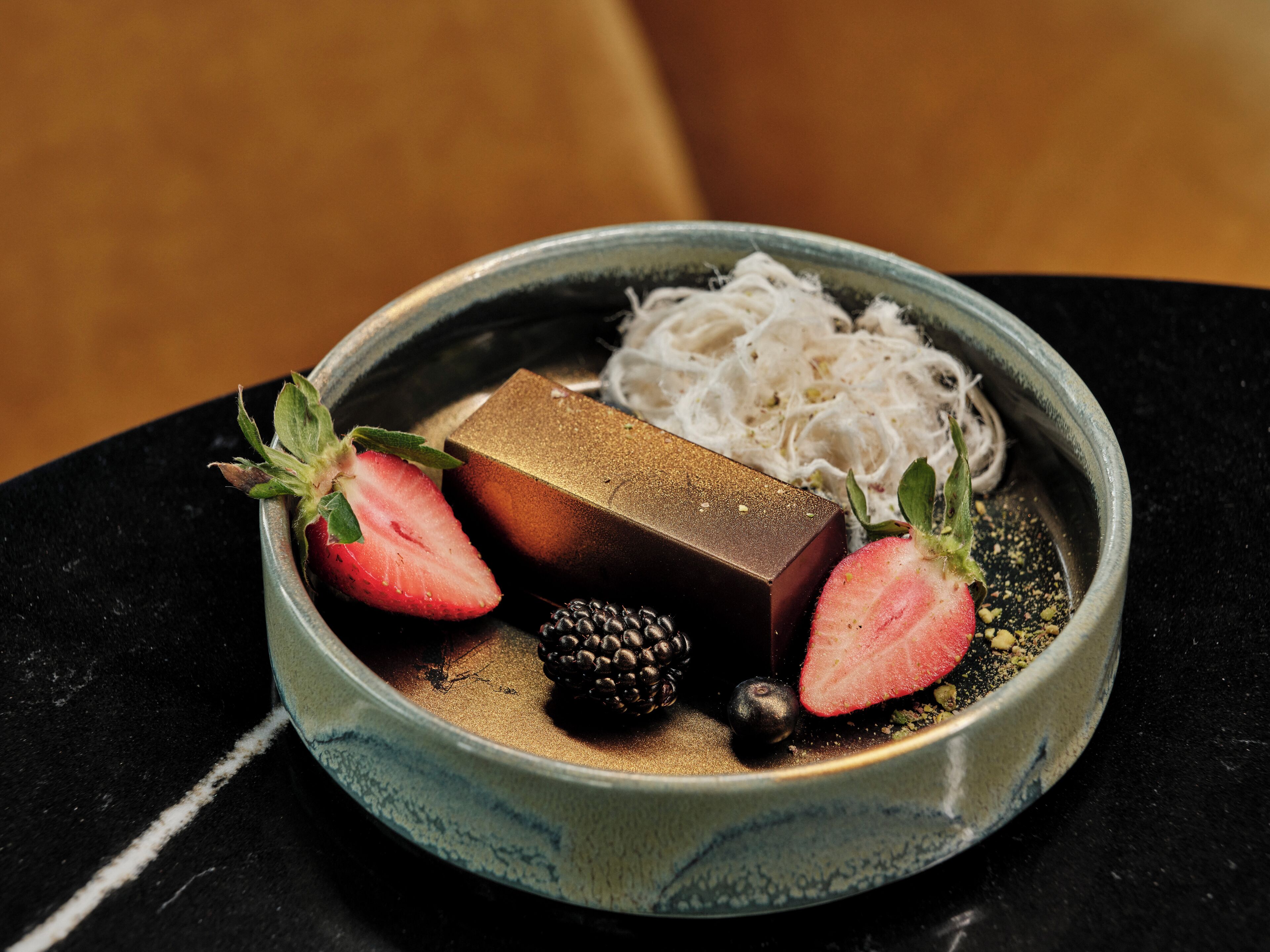 Uma tigela com sobremesa, morangos, um framboesa e um pedaço de chocolate, sobre superfície escura.