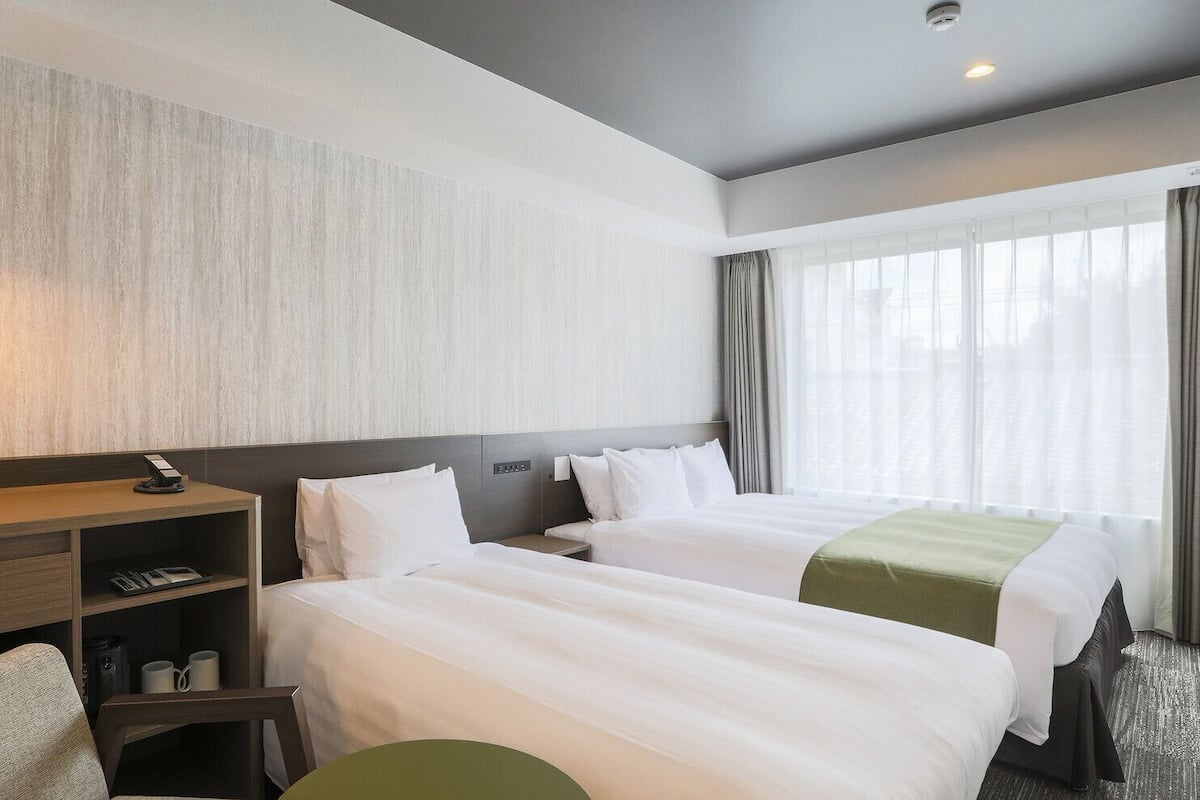 Quarto de hotel com duas camas, uma escrivaninha, uma cadeira e uma janela grande com cortinas.