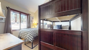 Quarto com cama, escrivaninha, mesa de cabeceira e janela com cortinas.