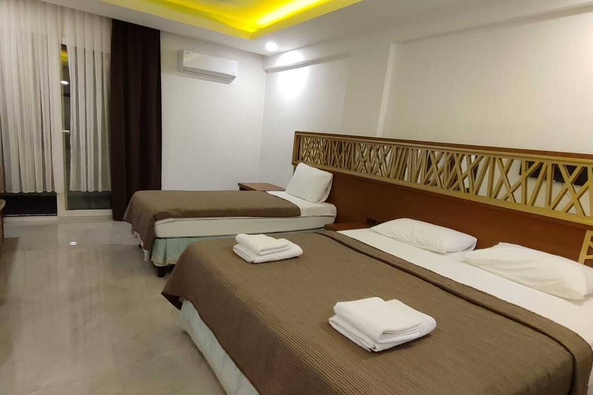 Quarto de hotel com três camas, cada uma com colcha marrom e almofadas brancas, e cabeceira com padrão geométrico.