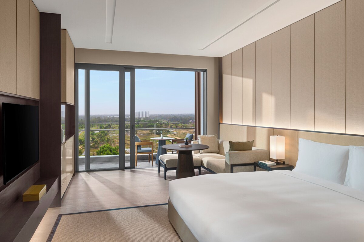 Quarto de hotel com uma cama grande, uma televisão, uma mesa com cadeira, uma mesa com cadeiras e vista para a paisagem urbana.