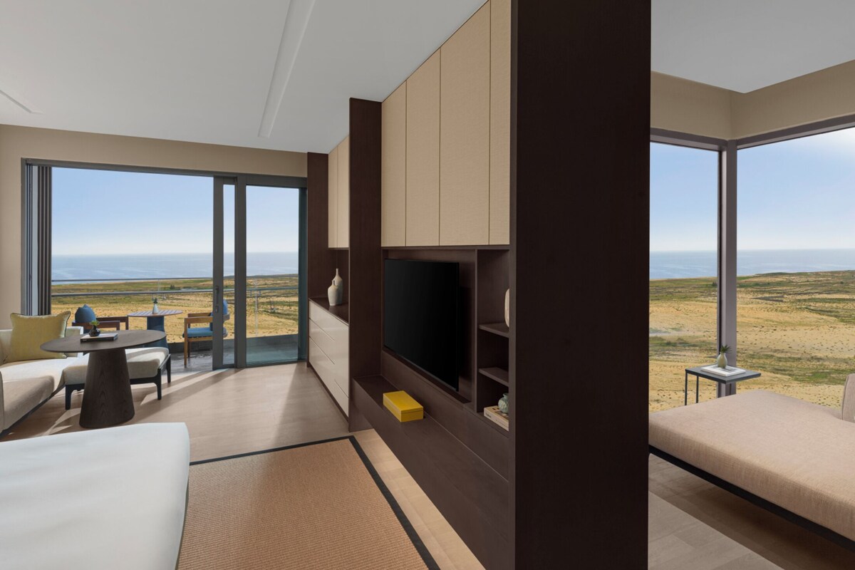 Quarto de hotel moderno com televisão grande, assentos confortáveis, sofá, área de jantar e varanda com vista para a praia.