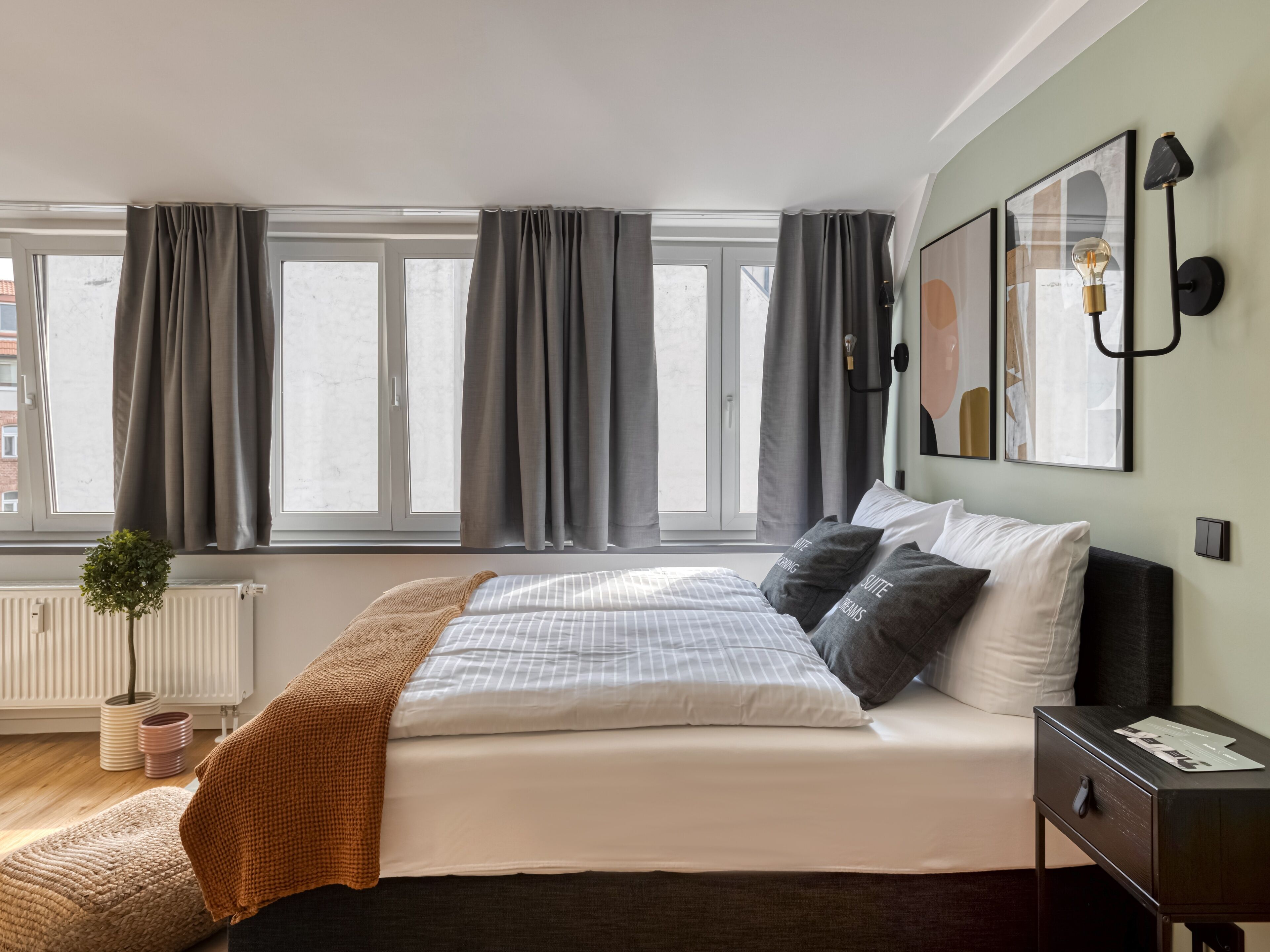 Quarto moderno com uma cama grande, cortinas cinzentas, um radiador, uma pequena planta e uma obra de arte na parede.
