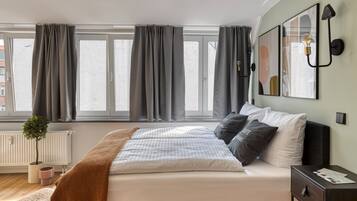 Quarto moderno com uma cama grande, cortinas cinzentas, um radiador, uma pequena planta e uma obra de arte na parede.