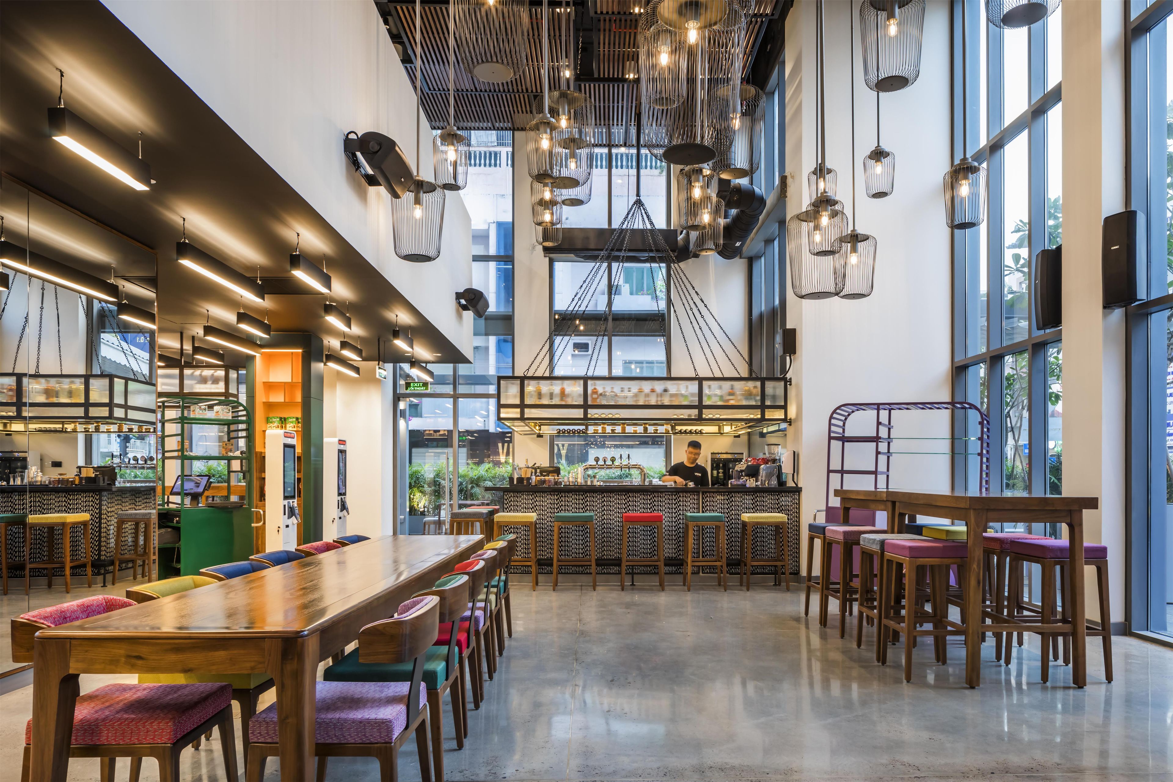 Um café amplo, com pé-direito alto, luminárias pendentes e grandes janelas.