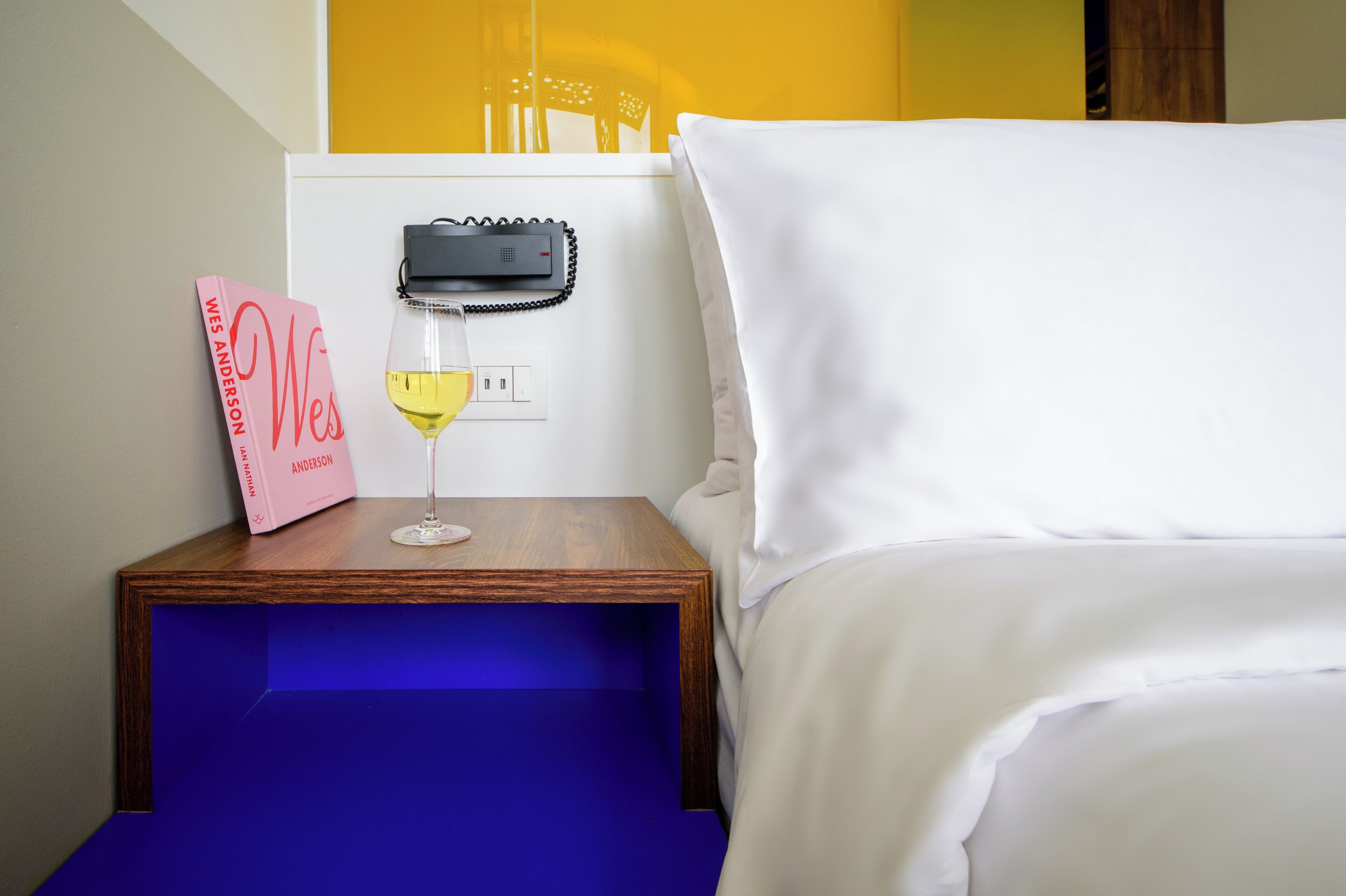 Quarto de hotel com uma cama, uma mesa de cabeceira com um livro, um copo de vinho e uma luminária.