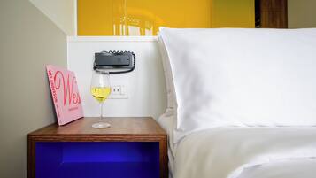 Quarto de hotel com uma cama, uma mesa de cabeceira com um livro, um copo de vinho e uma luminária.