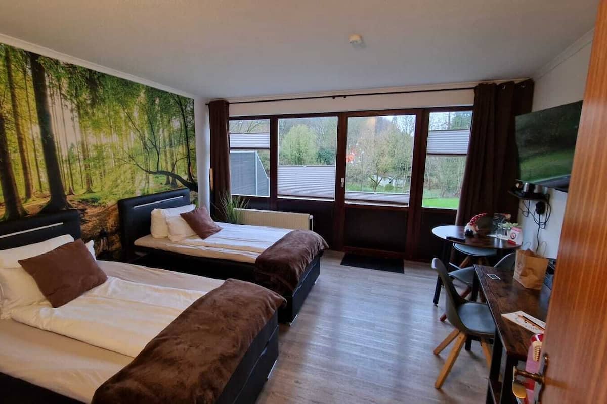 Quarto de hotel com duas camas, uma escrivaninha e uma janela grande com vista para árvores.