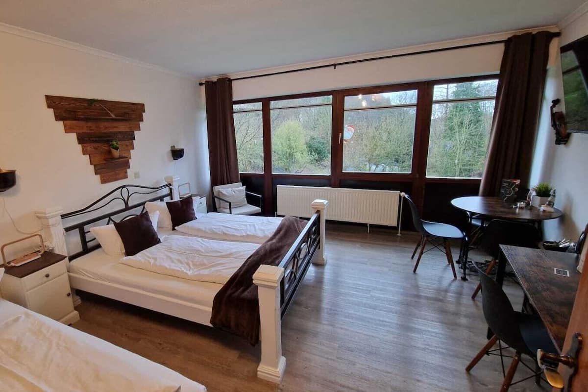 Quarto de hotel com duas camas de solteiro, uma mesinha com cadeiras, radiador e janelas amplas com cortinas.