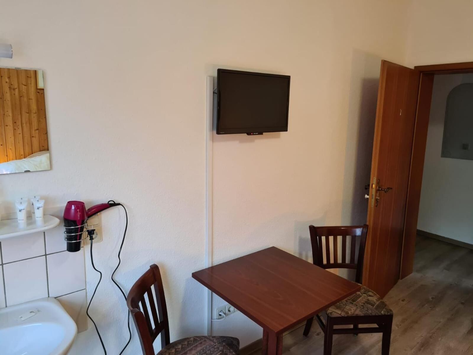Um quarto pequeno com uma mesa de madeira, duas cadeiras, uma televisão presa na parede e uma porta que dá para outro quarto.