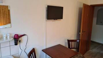 Um quarto pequeno com uma mesa de madeira, duas cadeiras, uma televisão presa na parede e uma porta que dá para outro quarto.
