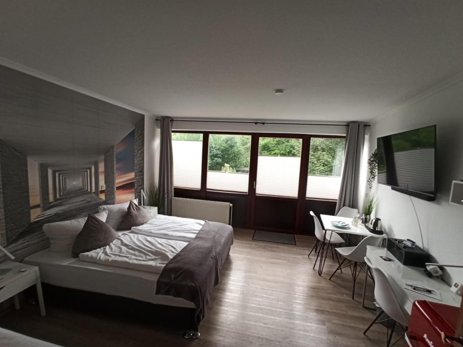 Quarto de hotel moderno com cama, escrivaninha e cadeira, janelas amplas e televisão na parede.