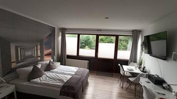 Quarto de hotel moderno com cama, escrivaninha e cadeira, janelas amplas e televisão na parede.
