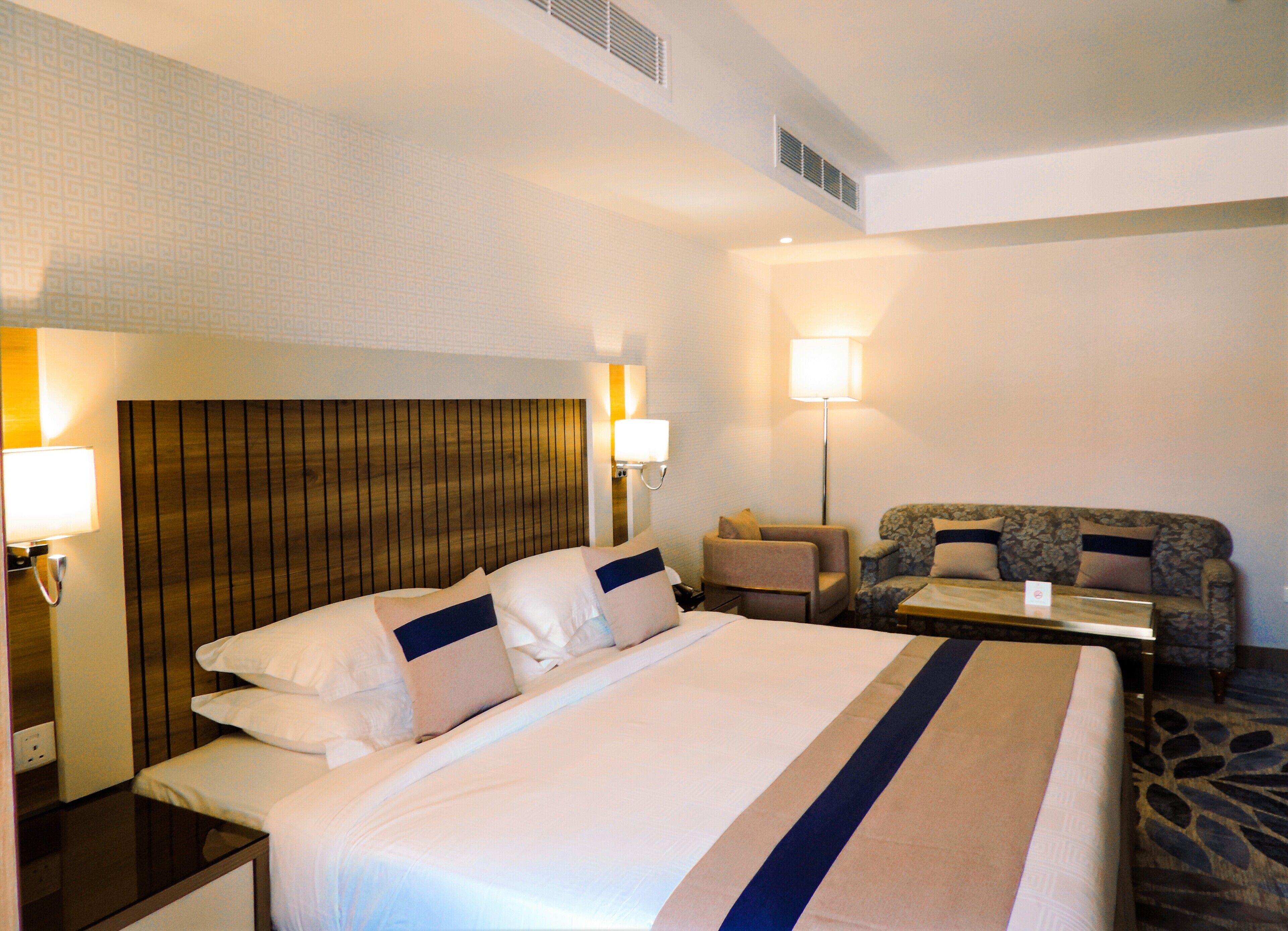 Quarto de hotel com uma cama grande, duas mesas de cabeceira, um sofá e uma luminária.
