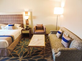Quarto de hotel com cama, sofá, poltrona, mesa pequena e mesa de cabeceira com luminária.