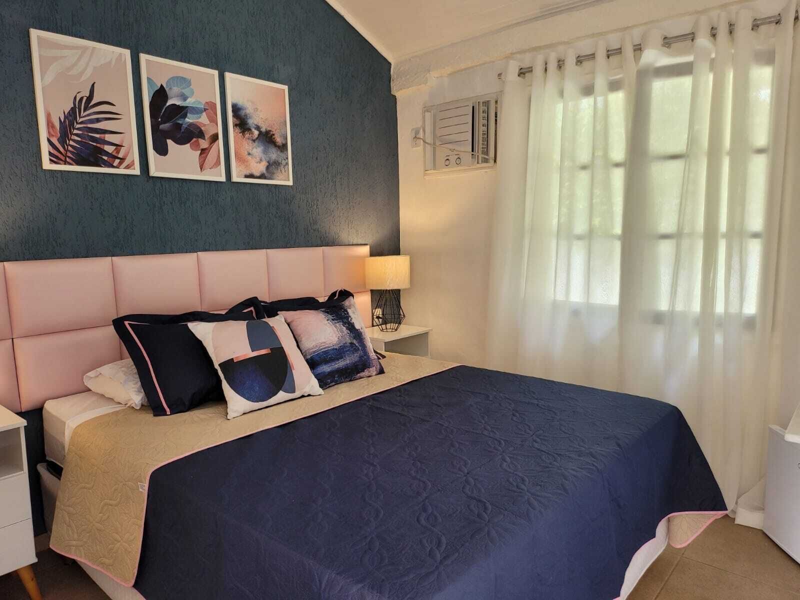 Quarto com cama, mesa de cabeceira, luminária e janela com cortinas.