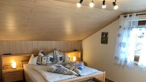 Quarto com teto inclinado, cabeceira de madeira, cama com roupa de cama estampada, abajures, cesto de vime e janela com cortinas finas.