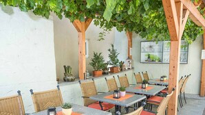 Uma área de refeições ao ar livre com várias mesas e cadeiras de vime, cercada por plantas em vasos e uma pérgola de madeira.