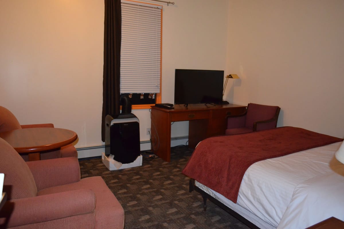 Quarto de hotel com cama, escrivaninha, cadeira, televisão e uma pequena mesa.