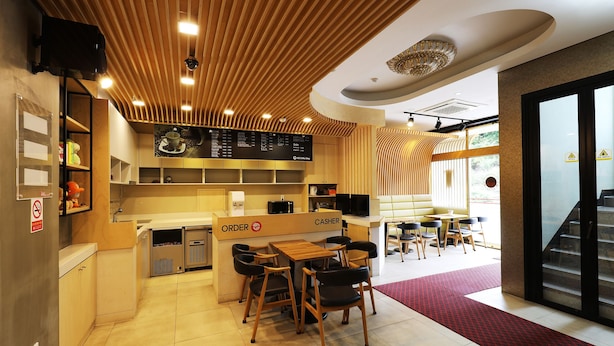 Um café moderno com painéis de madeira no teto, um balcão com placas 'ORDER' e 'CAIXA', e uma área de estar com mesas e cadeiras.
