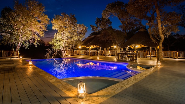 Uma área de piscina à noite com deck de madeira, espreguiçadeiras e estruturas com telhado de palha.