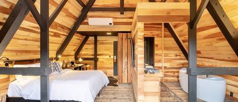 Quarto de sótão aconchegante, com teto de madeira, uma cama, uma escrivaninha e um banheiro pequeno.
