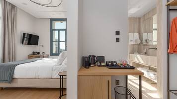 Quarto de hotel moderno com cama, escrivaninha com máquina de café e banheiro com espelho e porta-toalhas.
