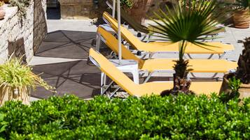 Uma área de pátio com cadeiras de lounge amarelas e um guarda-sol amarelo, cercada por plantas em vasos e uma parede de pedra.