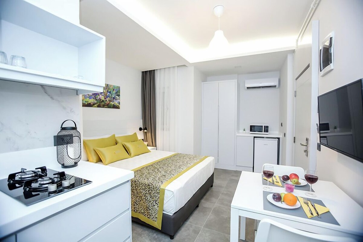 Quarto de hotel compacto com cozinha compacta, uma cama com almofadas amarelas, uma mesa de jantar com frutas e vinho, e uma televisão.