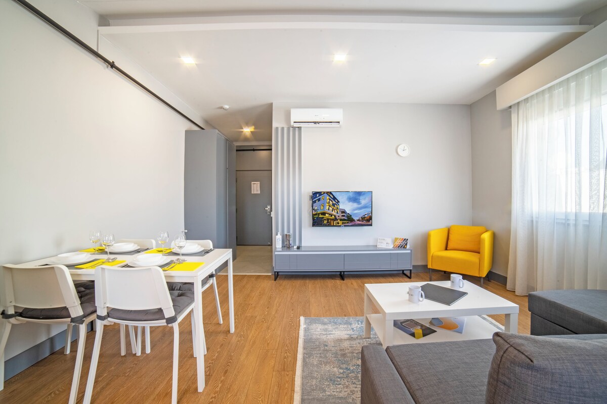 Uma sala de estar moderna com área de jantar, um sofá, uma poltrona amarela e um quadro na parede.