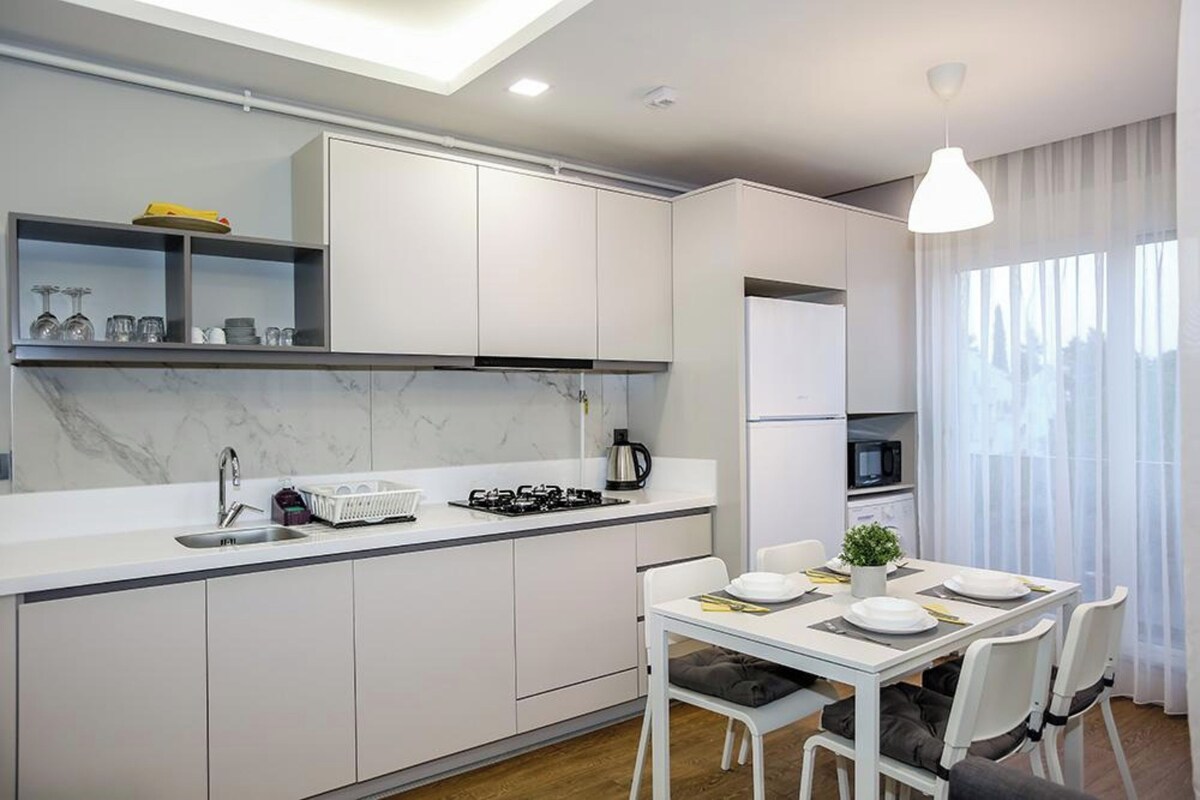 Uma cozinha moderna com armários brancos, painel de fundo de mármore, uma área de refeições com mesa e cadeiras, e uma luminária pendente.