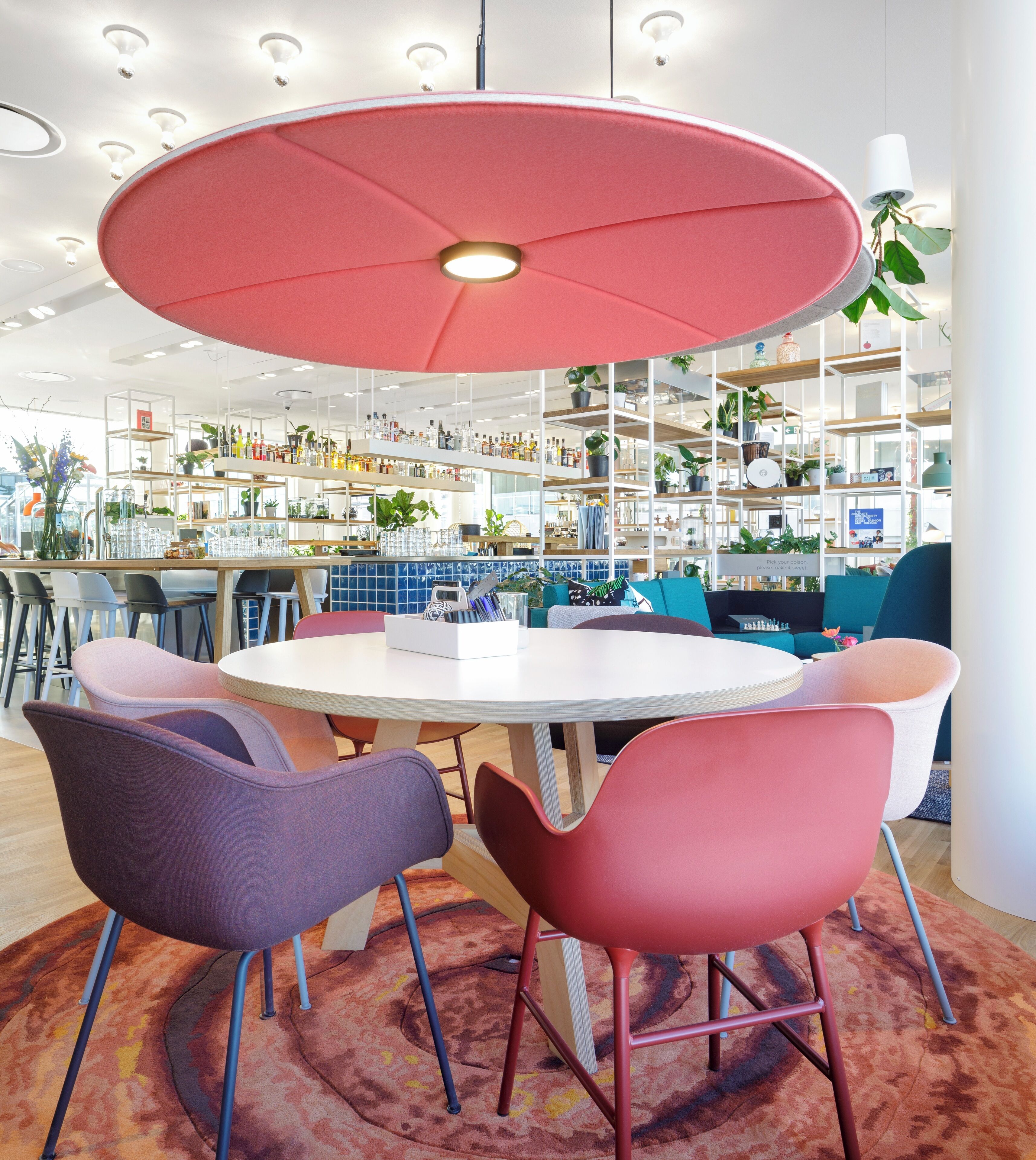 Um interior moderno com uma mesa redonda cercada por cadeiras coloridas, uma luminária vermelha no teto e uma planta à direita.