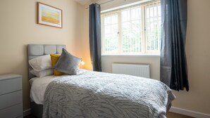 Quarto com cama, mesa de cabeceira, radiador, janela com cortinas e um quadro na parede.