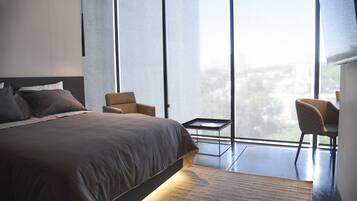 Quarto moderno com uma cama grande, duas cadeiras, uma mesa pequena e vista para a cidade através de grandes janelas.