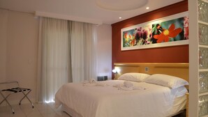 Quarto de hotel com uma cama grande, um quadro de flores na parede, uma janela com cortinas e um radiador.