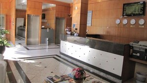 Uma área de recepção com piso de mármore, painéis de madeira, um balcão de recepção, um relógio e uma tela de televisão.