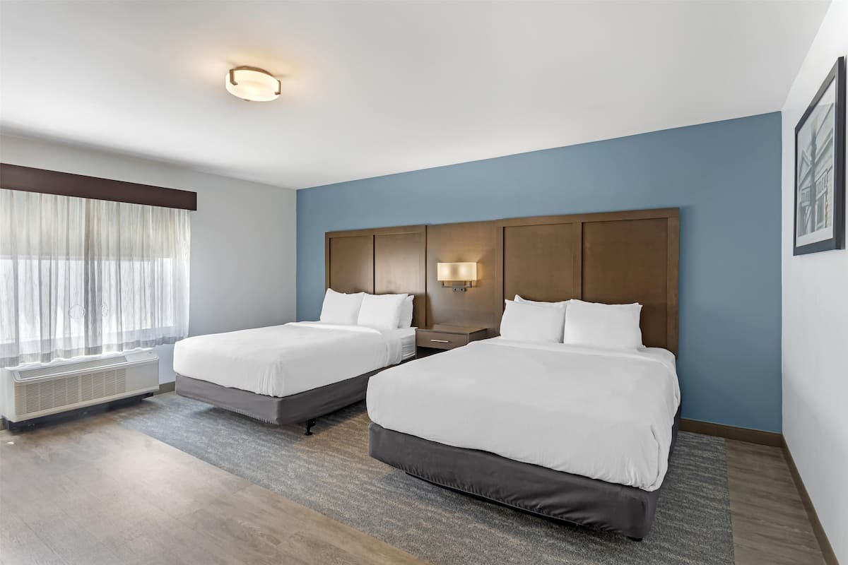 Quarto de hotel com duas camas, uma janela grande com cortinas leves, uma luminária na parede e um lustre no teto.