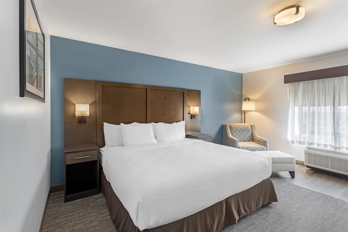 Quarto de hotel com cama grande, mesas de cabeceira, uma cadeira, janela com cortinas e luminárias na parede.