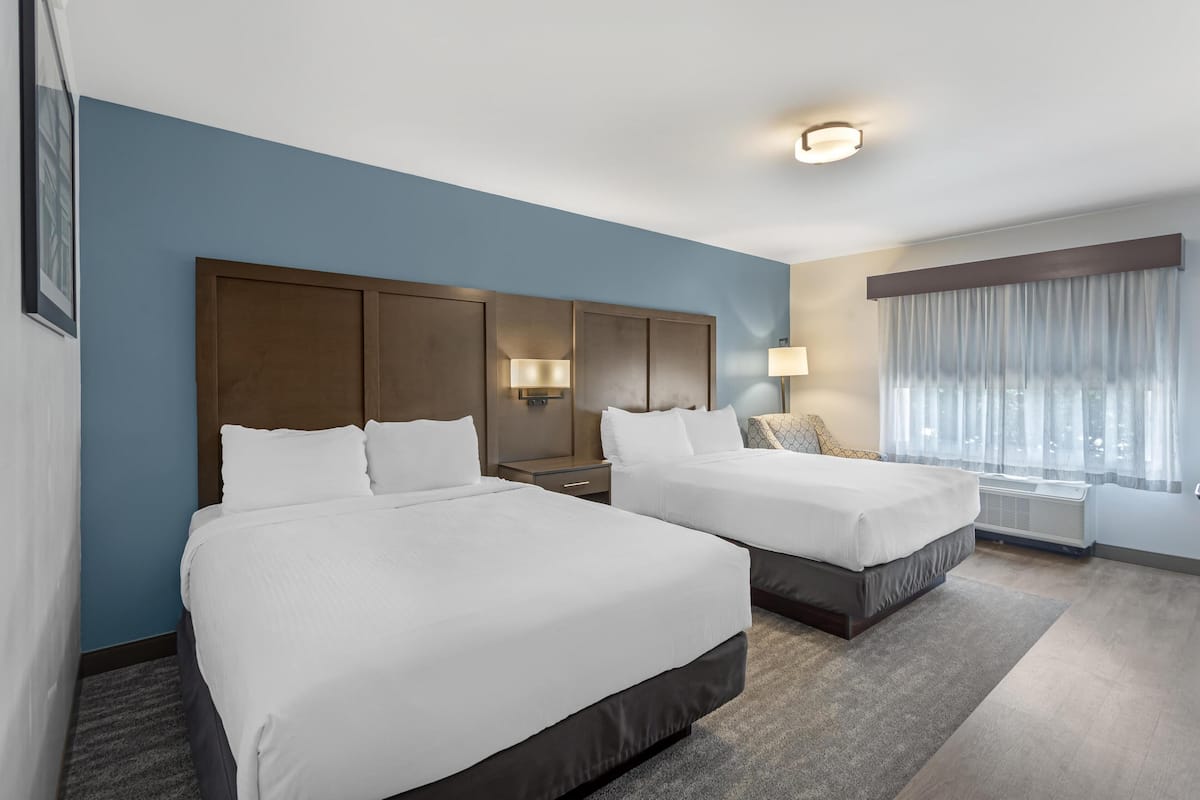 Quarto de hotel com duas camas, uma janela grande com cortinas, uma luminária na parede e uma luz no teto.