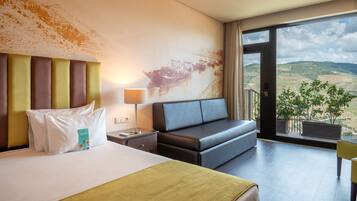 Quarto de hotel com uma cama grande, um sofá, uma mesa de cabeceira, um abajur e vista para o campo através de uma grande janela.
