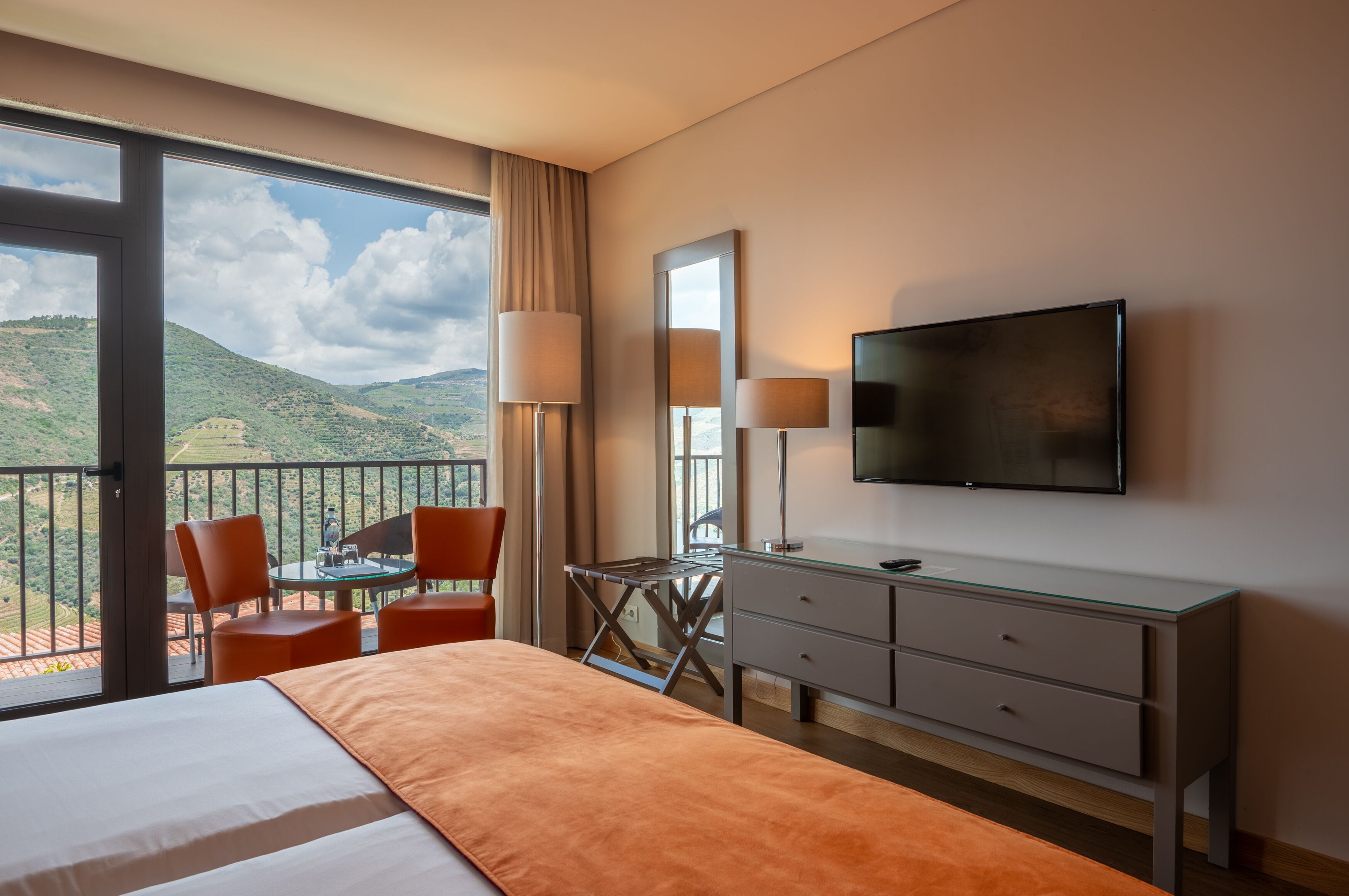 Quarto de hotel com cama, escrivaninha, cadeira, televisão e varanda com vista para as montanhas.