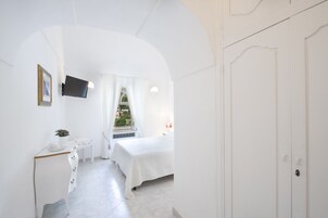 Quarto branco com uma cama, uma escrivaninha, uma cadeira, uma TV na parede e uma janela com cortinas.