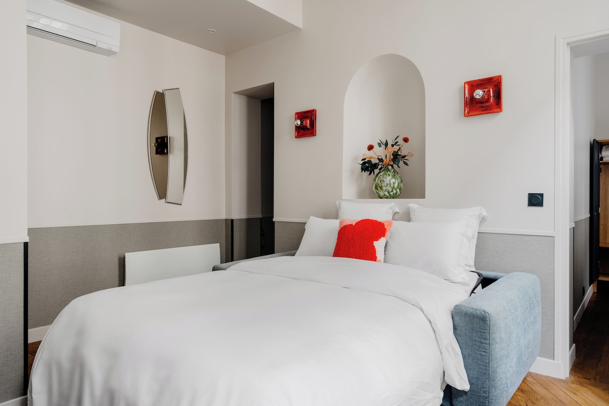 Quarto de hotel com uma cama grande, um espelho, um vaso com flores e um interruptor de luz vermelho.